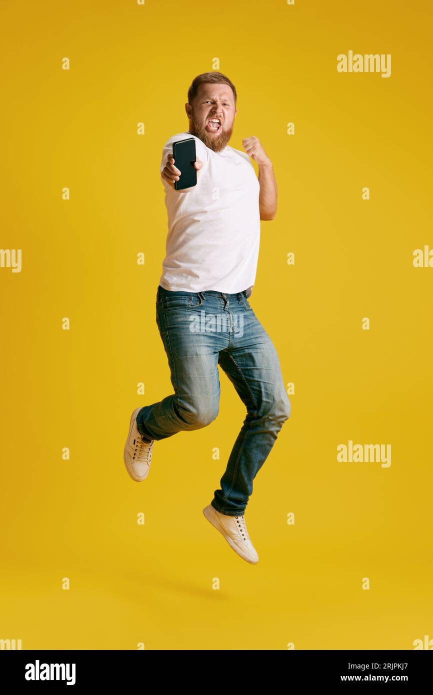 Man emotionally jumping with mobile phone against yellow studio ...