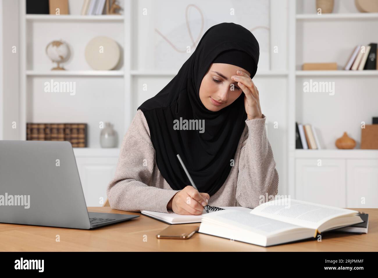 Tired Muslim woman in hijab working near laptop at wooden table in room ...