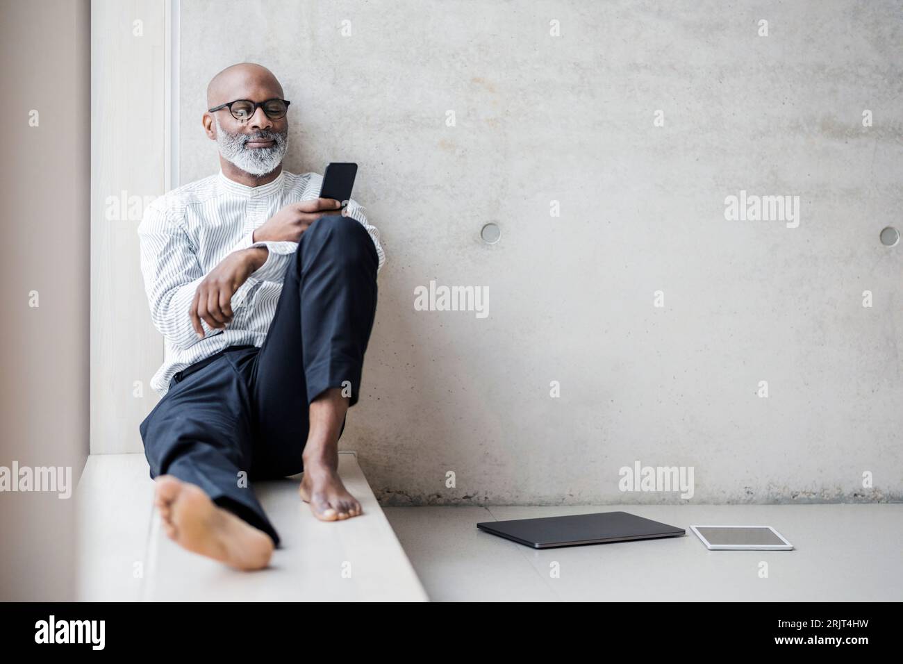 Barefoot mature businessman sitting on window sill looking at ...