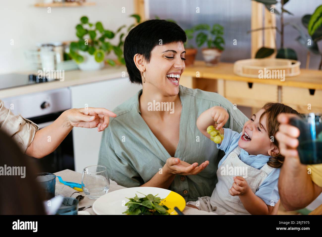 Smiling son holding grapes sitting on mother's lap at dining table in ...