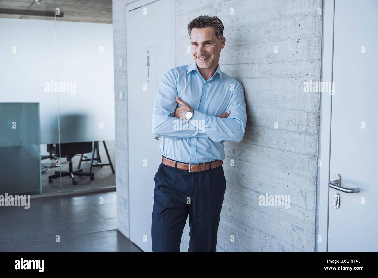 Happy man with arms crossed leaning on wall in office Stock Photo - Alamy