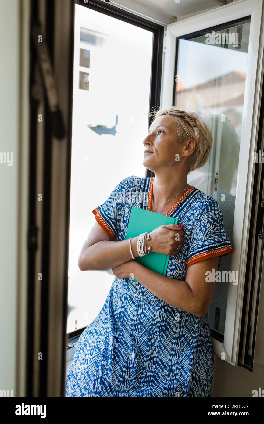 Contemplative mature woman with book looking through window Stock Photo ...