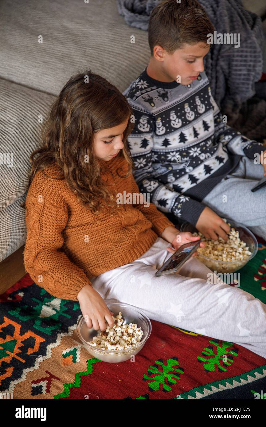 Siblings using smart phones and eating popcorn at home Stock Photo - Alamy
