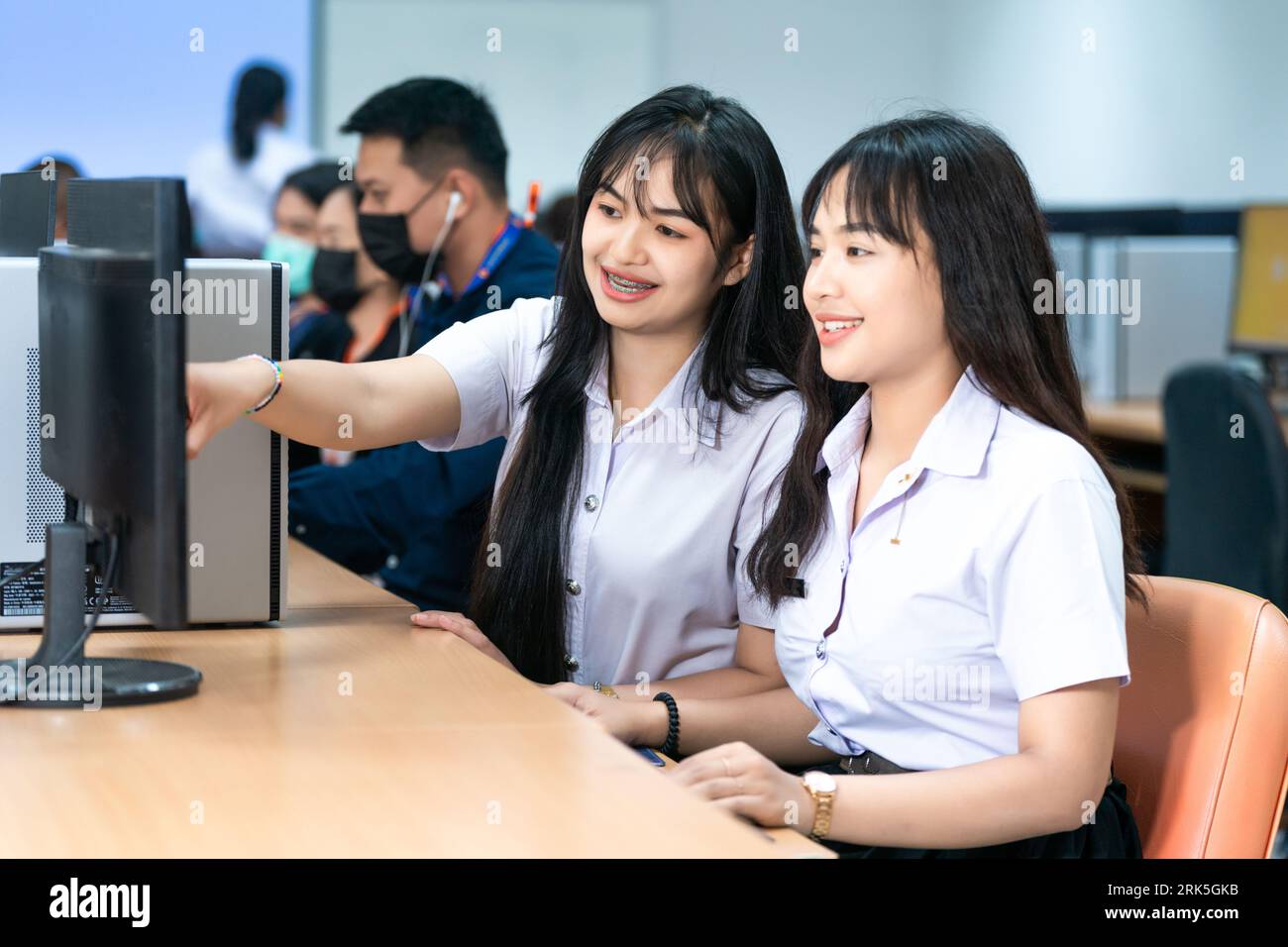 Two young Asian female students are seated at a desk, interacting with ...