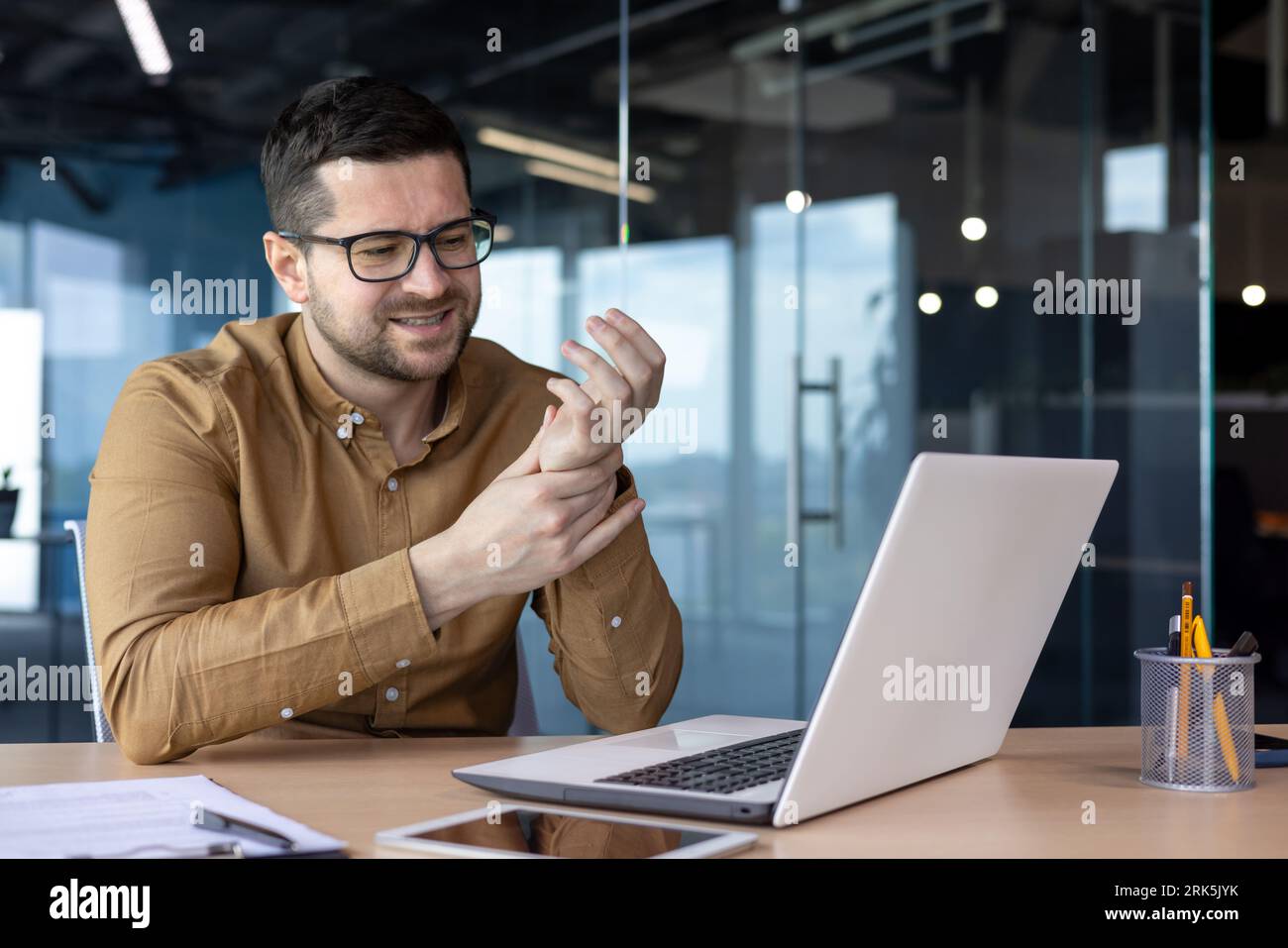 Tired young man sitting at the desk in the office and holding his wrist ...