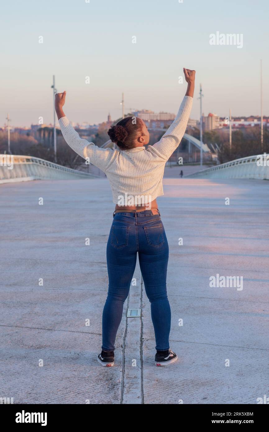 A mid-adult woman wearing blue jeans is joyfully raising her arms in ...