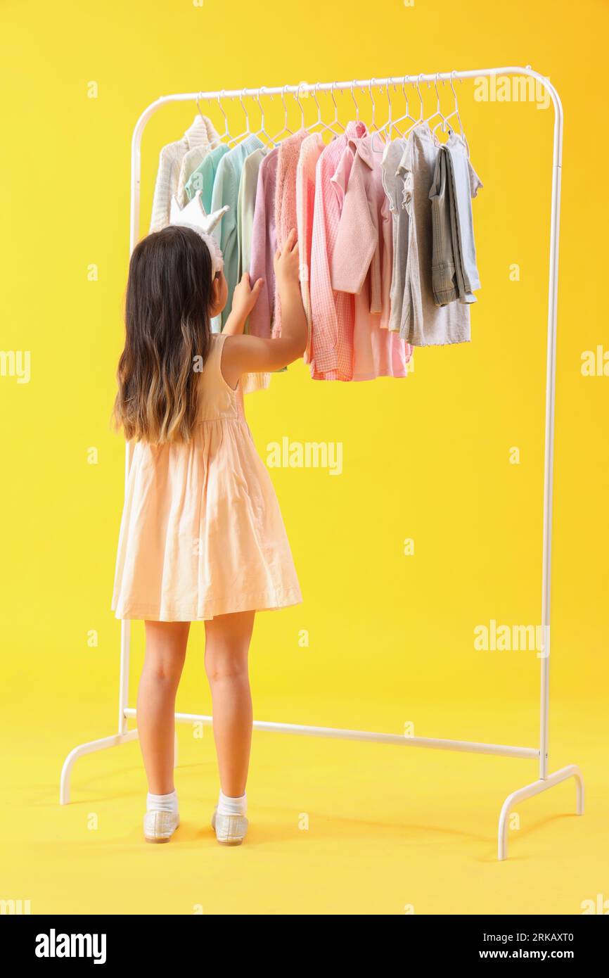 Cute little girl near rack with stylish clothes on yellow background ...