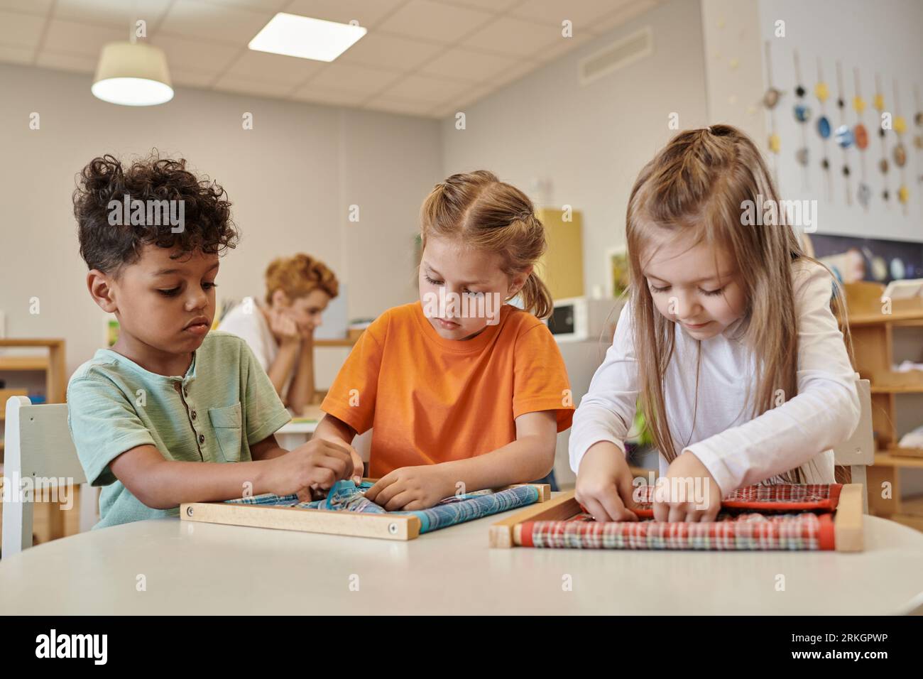 multiethnic kids playing with cloth and buttons on table in montessori ...