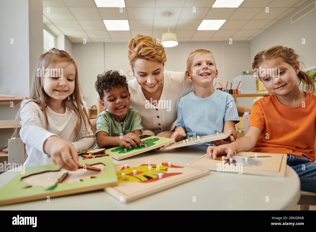 cheerful teacher sitting near multiethnic kids playing with didactic ...
