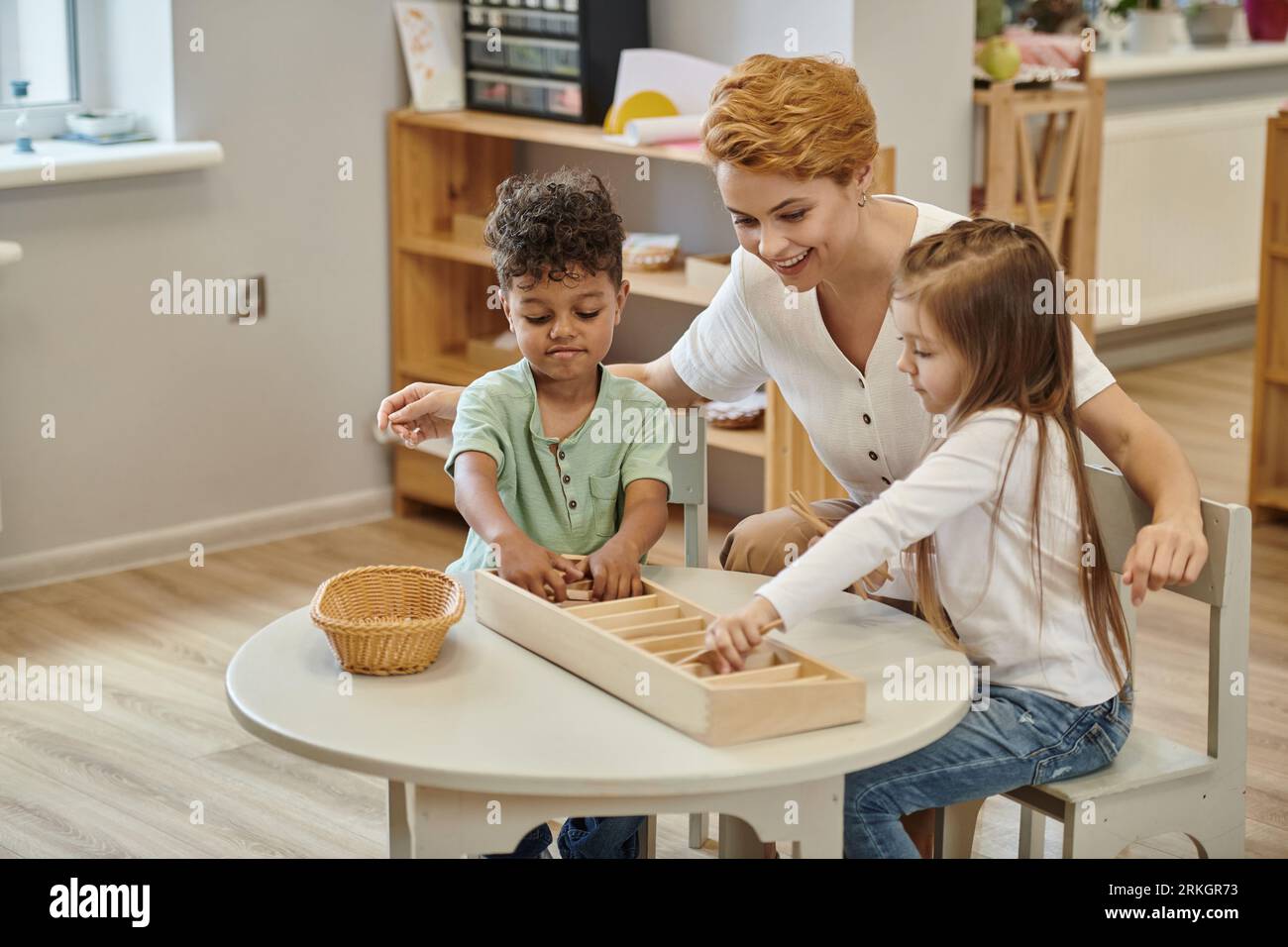 happy teacher talking to interracial kids playing with wooden didactic ...