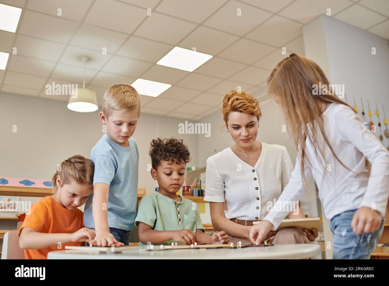 teacher sitting near multiethnic kids with didactic material playing in ...