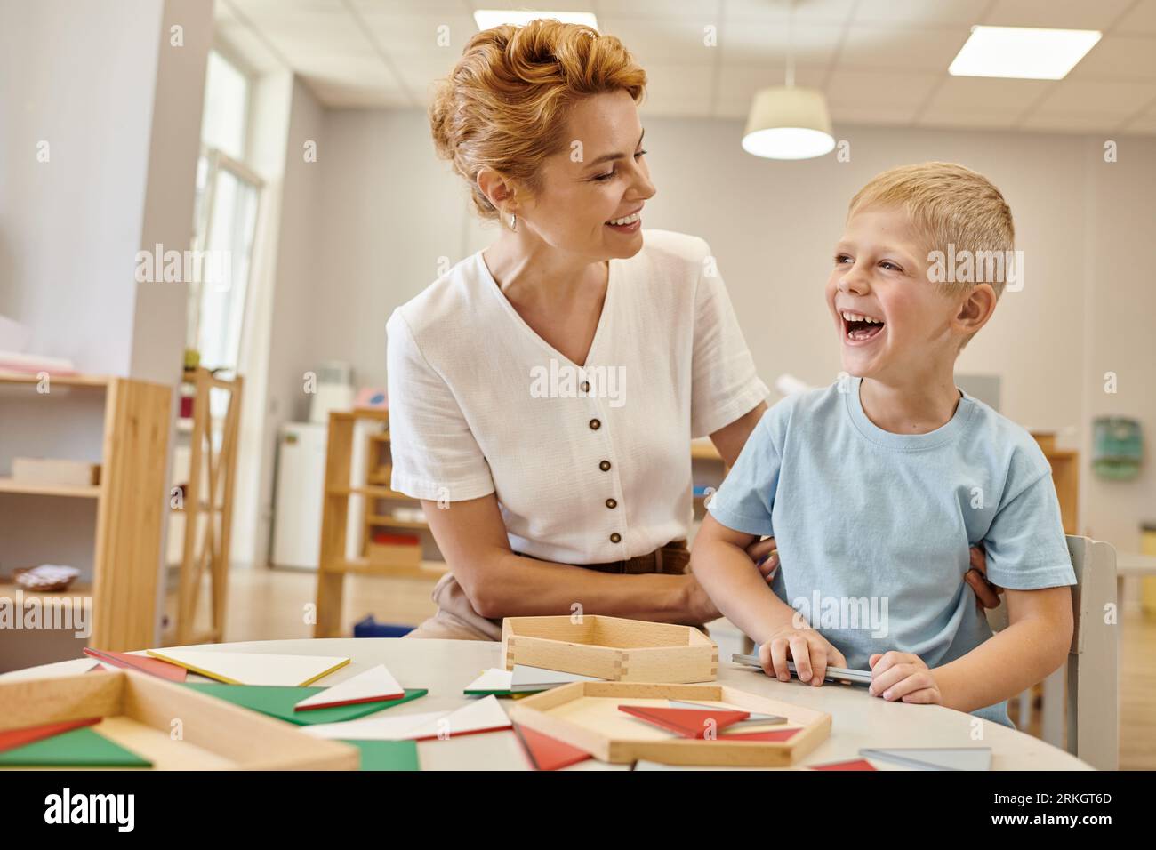carefree teacher hugging boy near wooden game on table in montessori ...