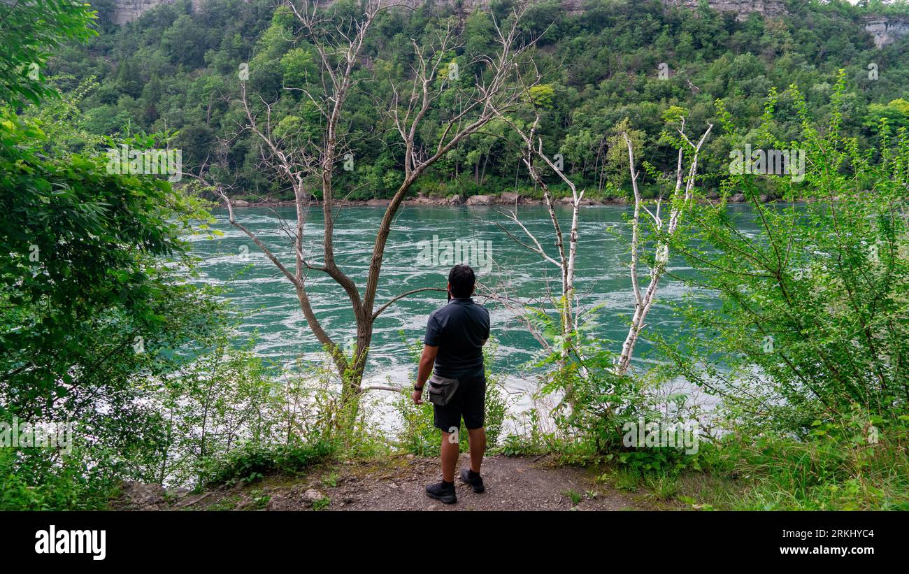 An adult male with his back to the camera, gazing out at a river in the ...
