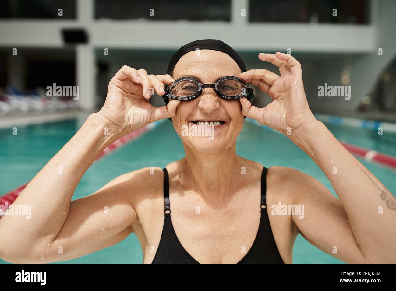 happy and tattooed woman in swim cap wearing goggles, positive ...