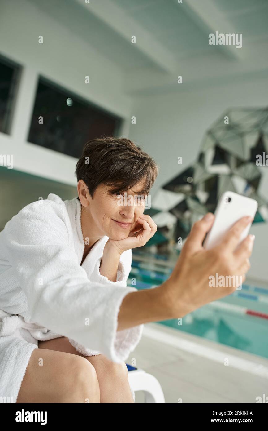 positive middle aged woman in white robe taking selfie, smartphone, spa center, swimming pool ...