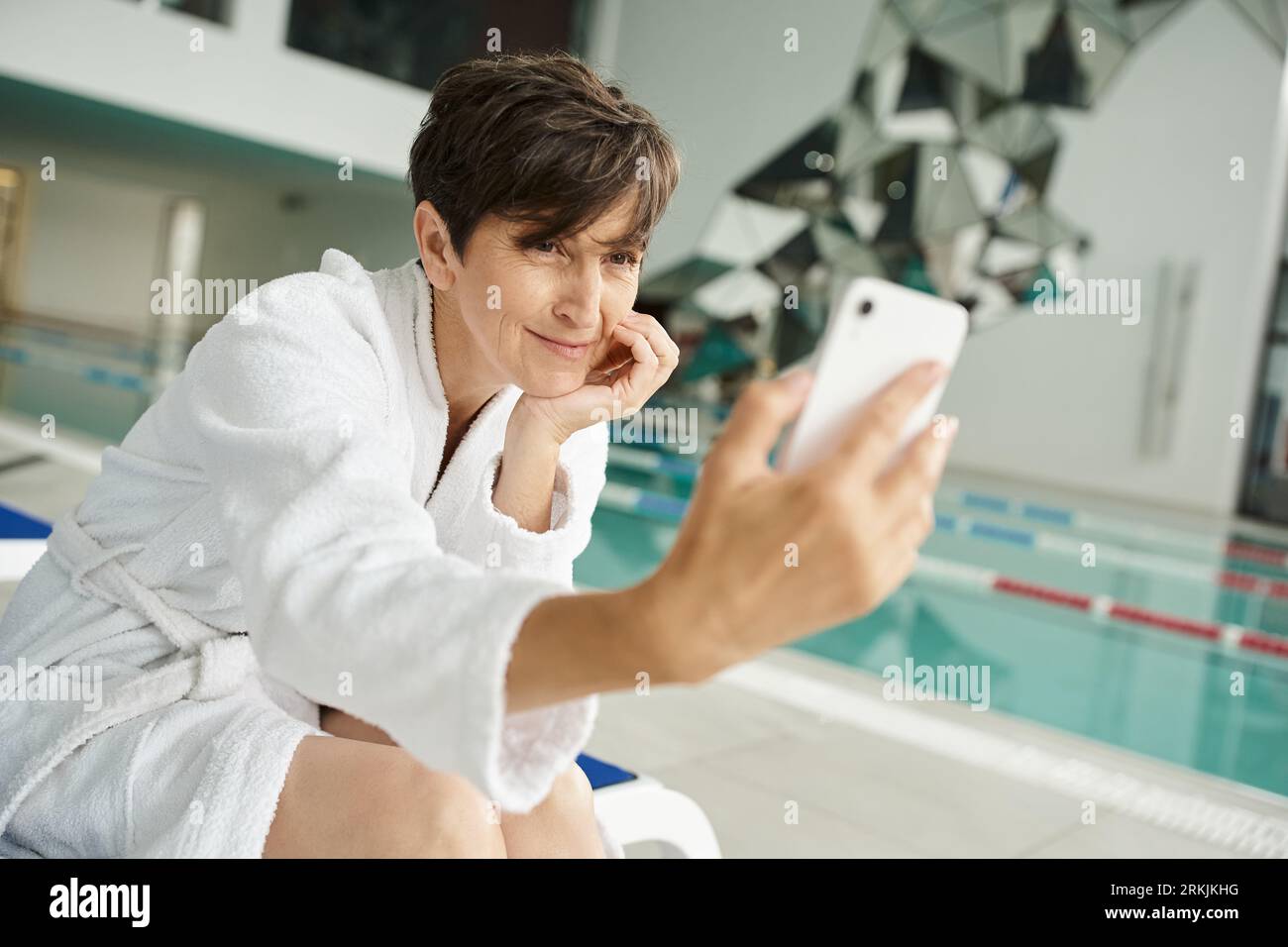 cheerful middle aged woman in white robe taking selfie, smartphone, spa center, swimming pool ...