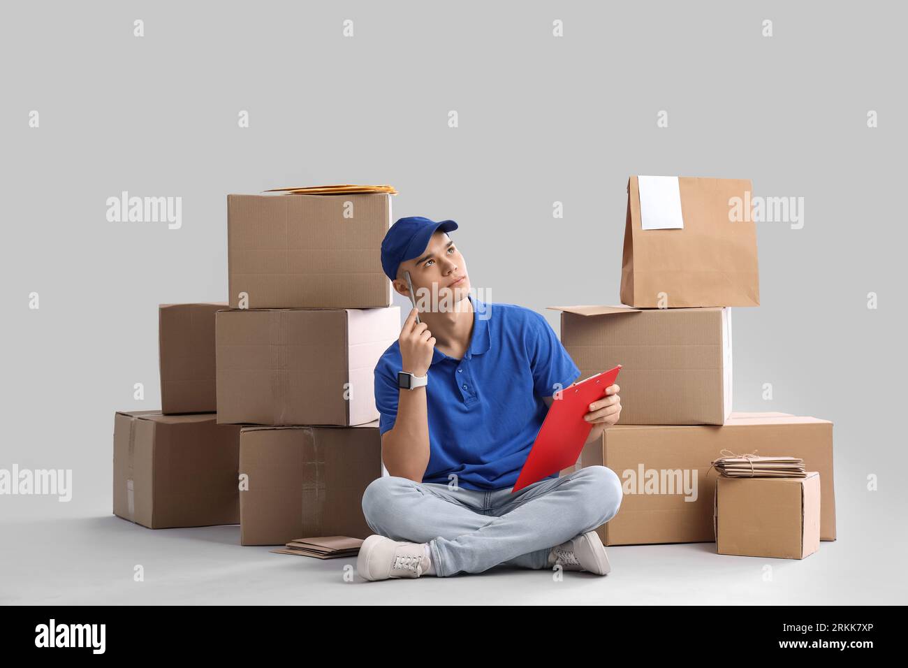 Thoughtful young courier with clipboard and parcels on light background ...