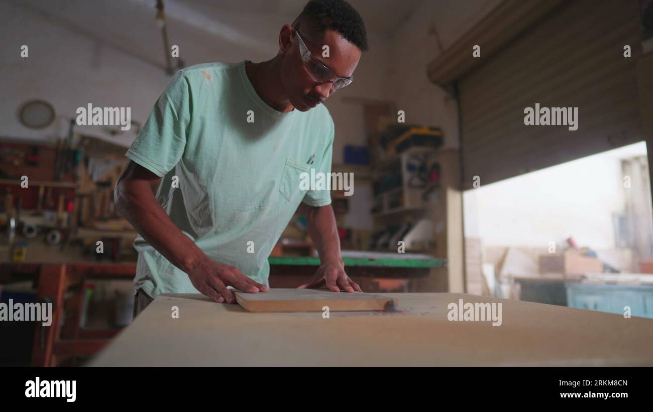 One young black carpenter using machine to trim piece of wood at ...