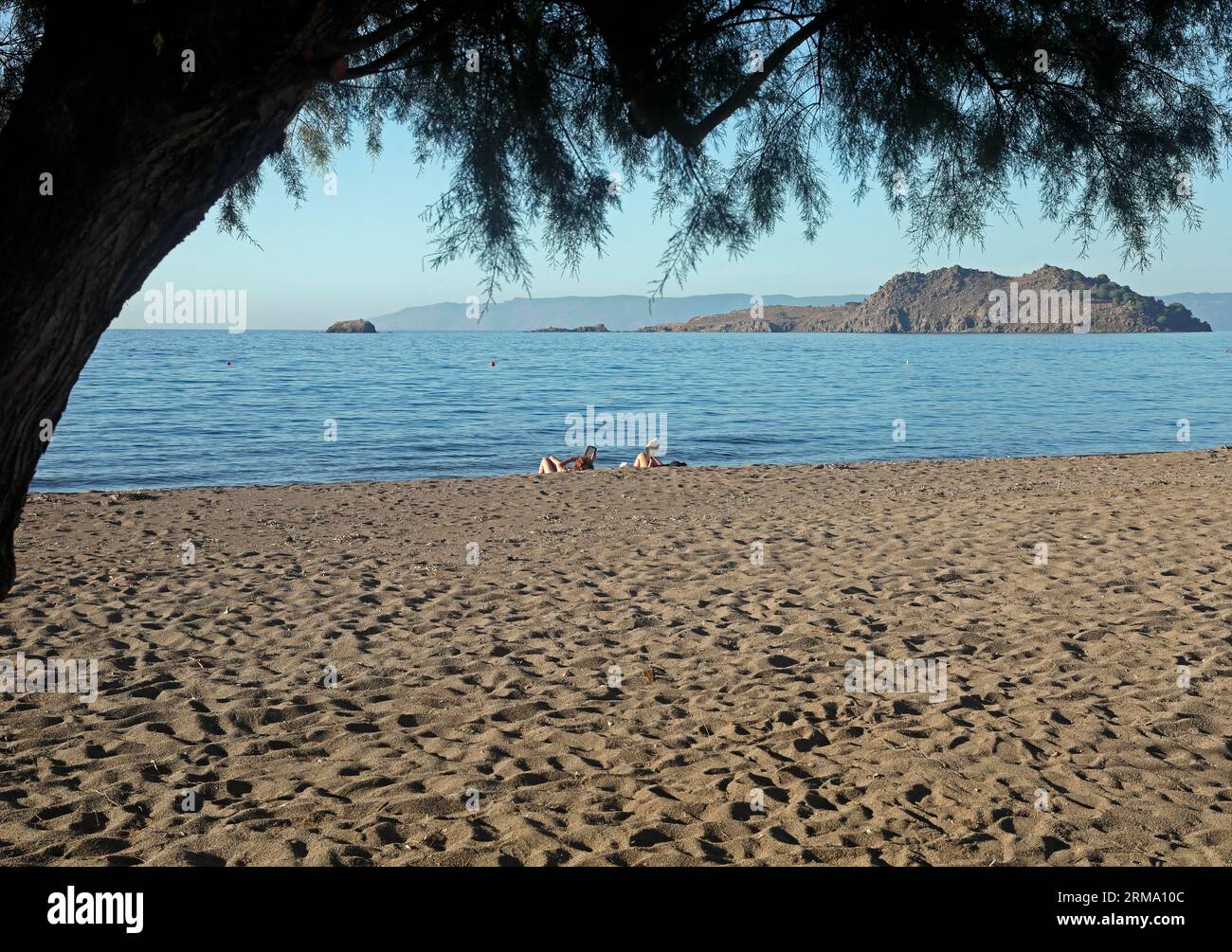 Two women reading, relaxing and sunbathing on a sandy beach, Lesbos scenes, Greece. Taken 2022 ...