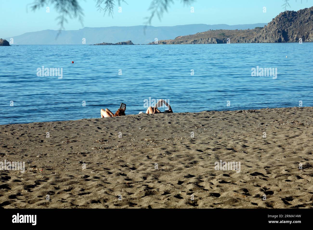Two women reading, relaxing and sunbathing on a sandy beach, Lesbos scenes, Greece. Taken 2022 ...