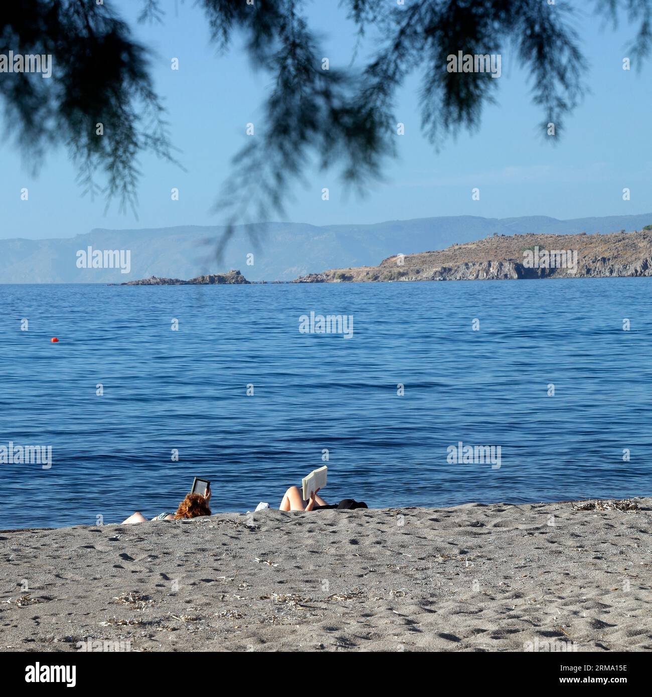 Two women reading, relaxing and sunbathing on a sandy beach, Lesbos scenes, Greece. Taken 2022 ...