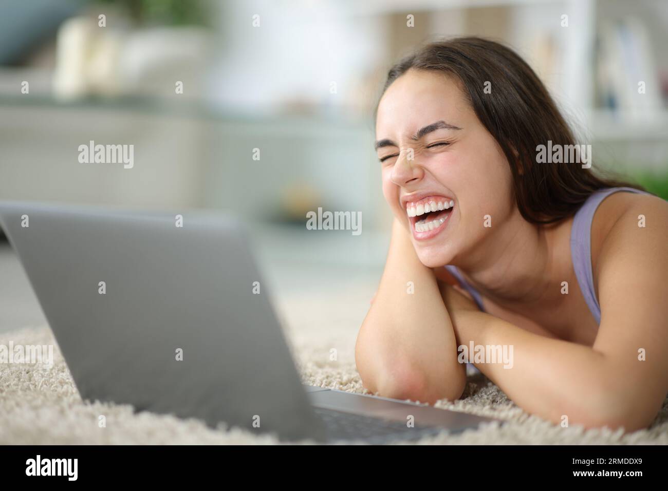 Happy woman laughing loudly watching media on laptop lying on the floor ...