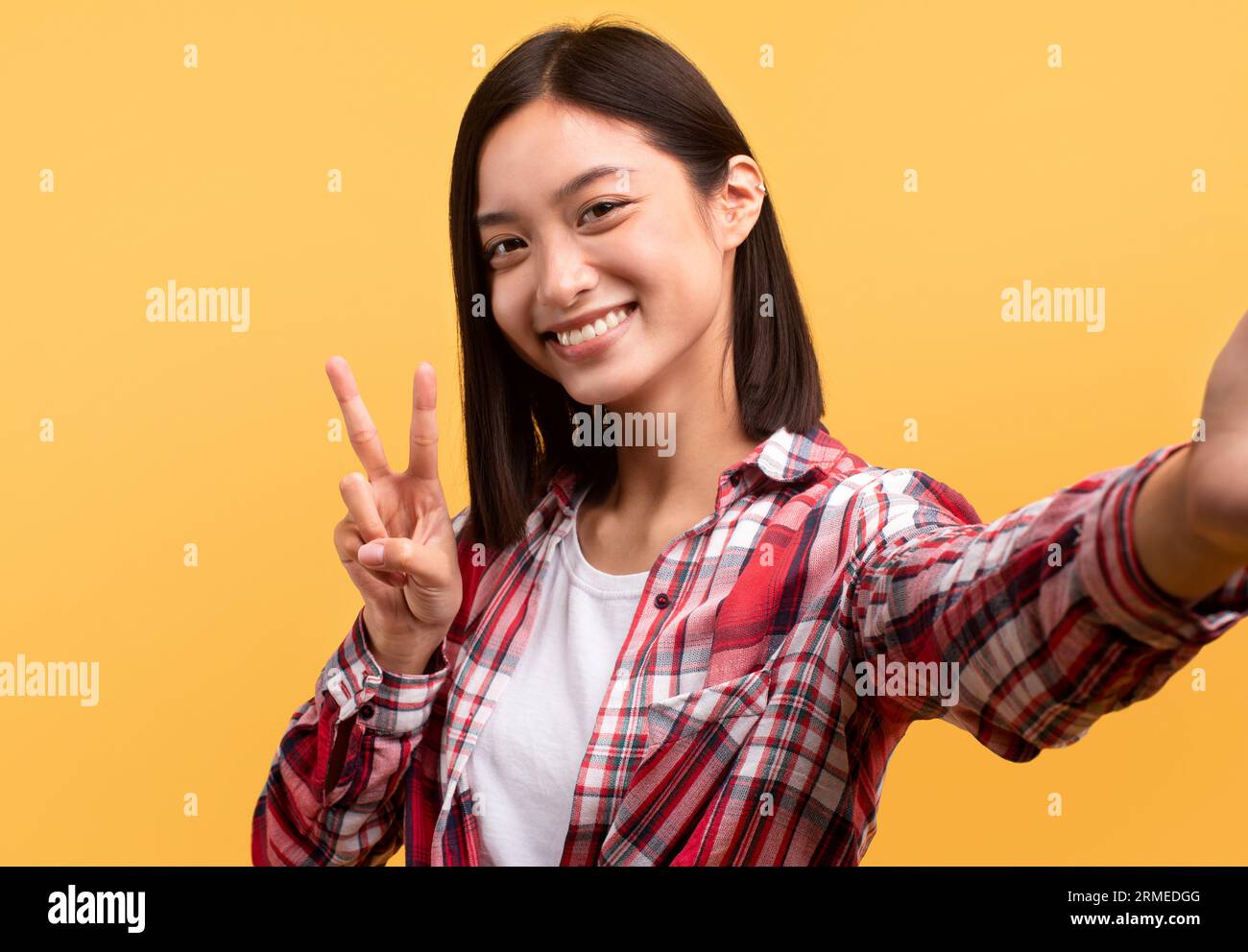 Glad japanese woman taking selfie and showing peace sign with hand posing isolated on yellow ...