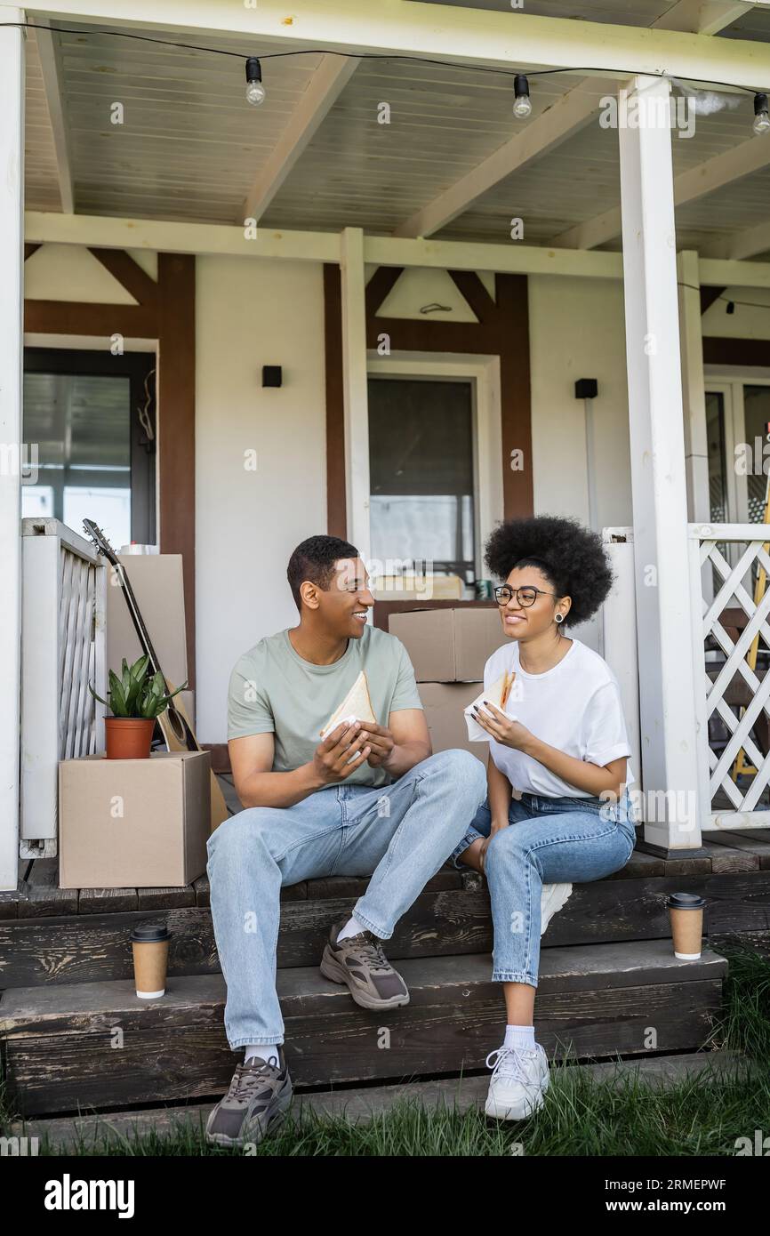 happy african american woman holding sandwich near boyfriend and coffee ...