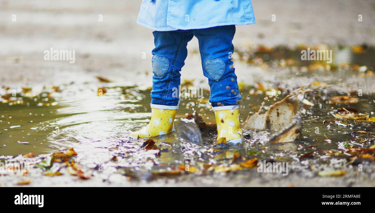 Child wearing yellow rain boots and jumping in puddle on a fall day. Toddler girl having fun ...