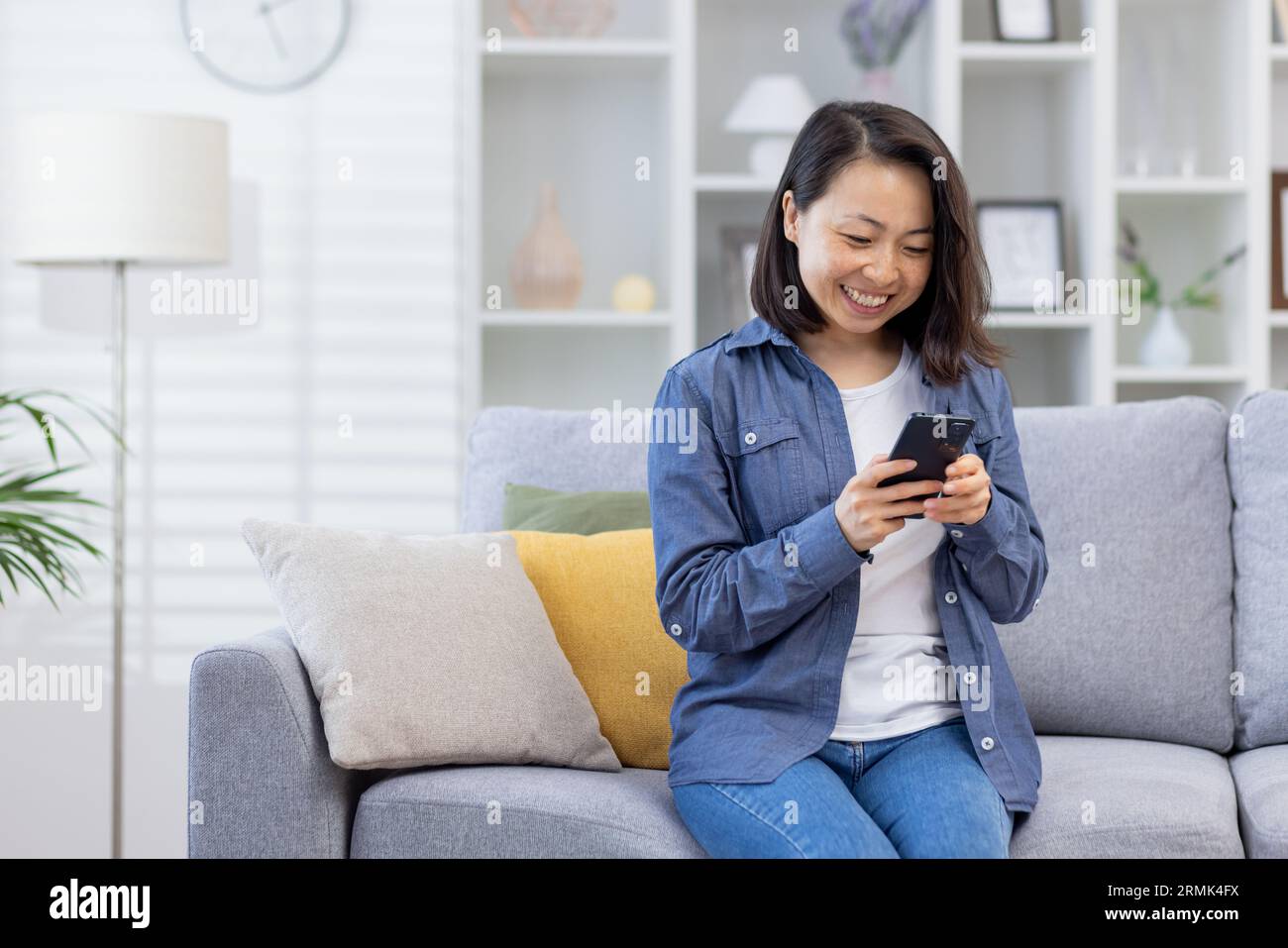 A young beautiful Asian sits alone at home on the sofa, the woman uses ...