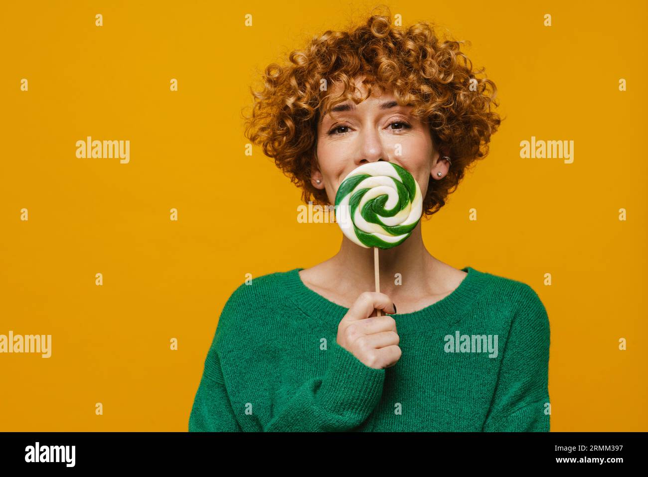 Curly ginger woman smiling while posing with lollipop isolated over ...