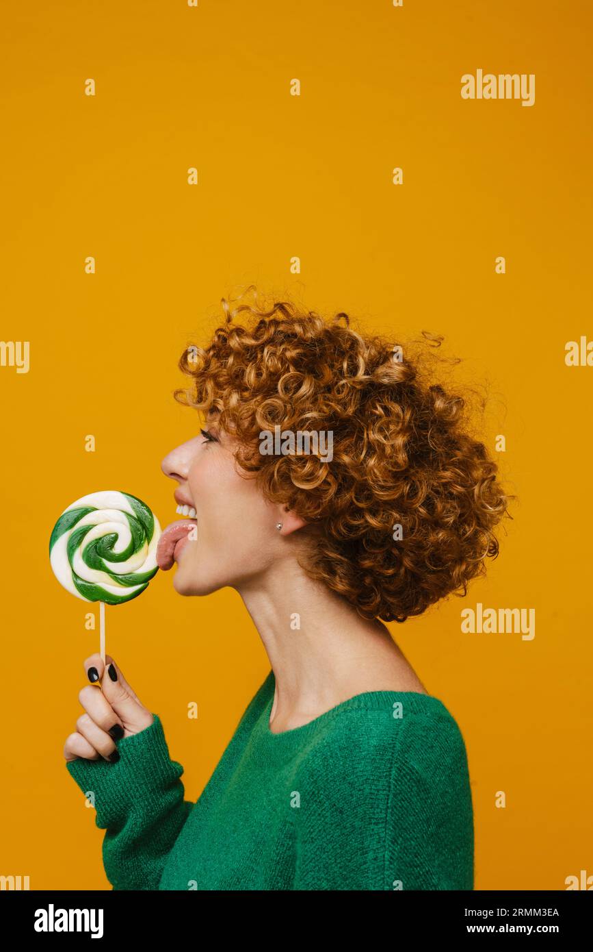 Middle-aged ginger smiling woman liking lollipop isolated over yellow ...