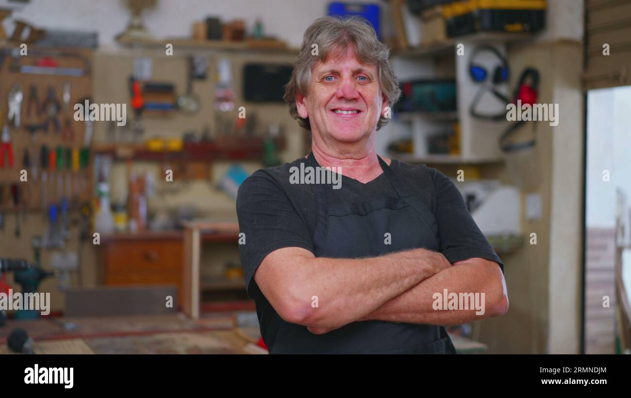 Portrait of a happy master carpenter standing inside workshop with arms ...