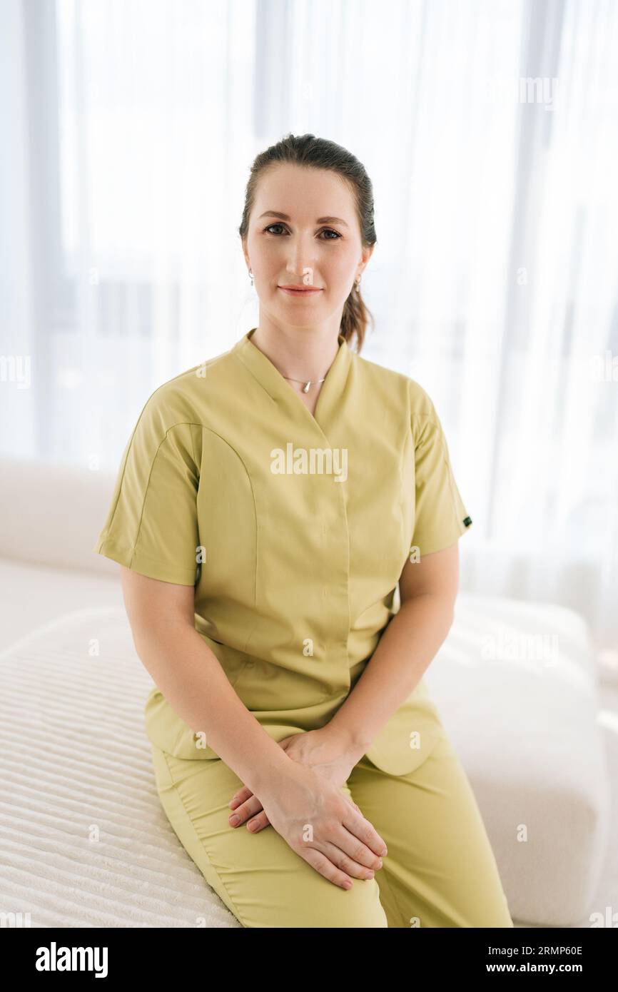 Vertical portrait of professional friendly female masseuse doctor in uniform sitting posing on ...