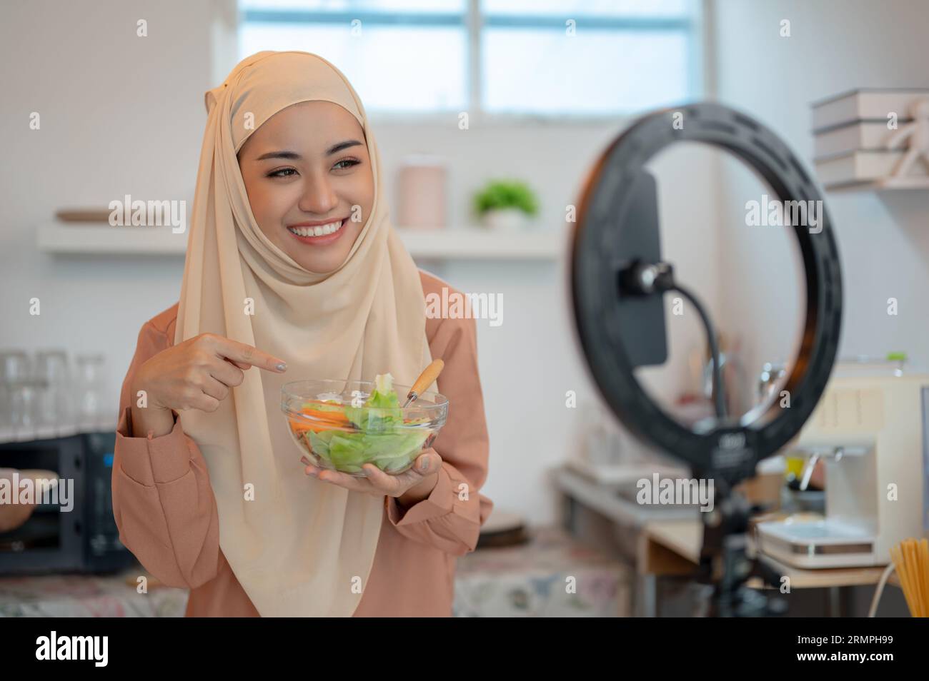 A beautiful Asian Muslim female influencer or food blogger is in the kitchen, live streaming her ...