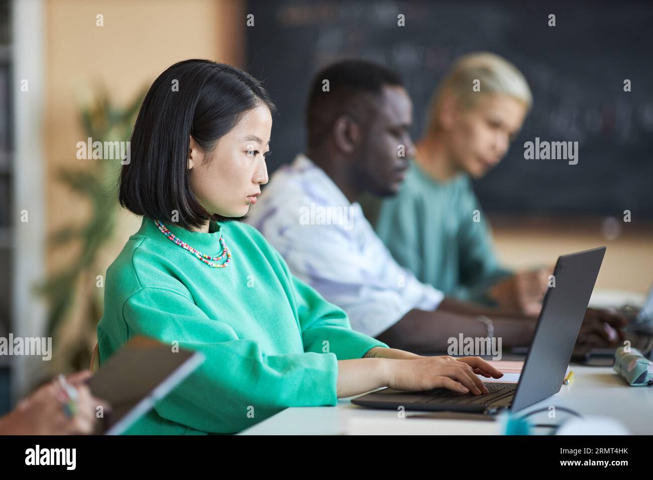Young serious Asian female student in green pullover looking at laptop ...