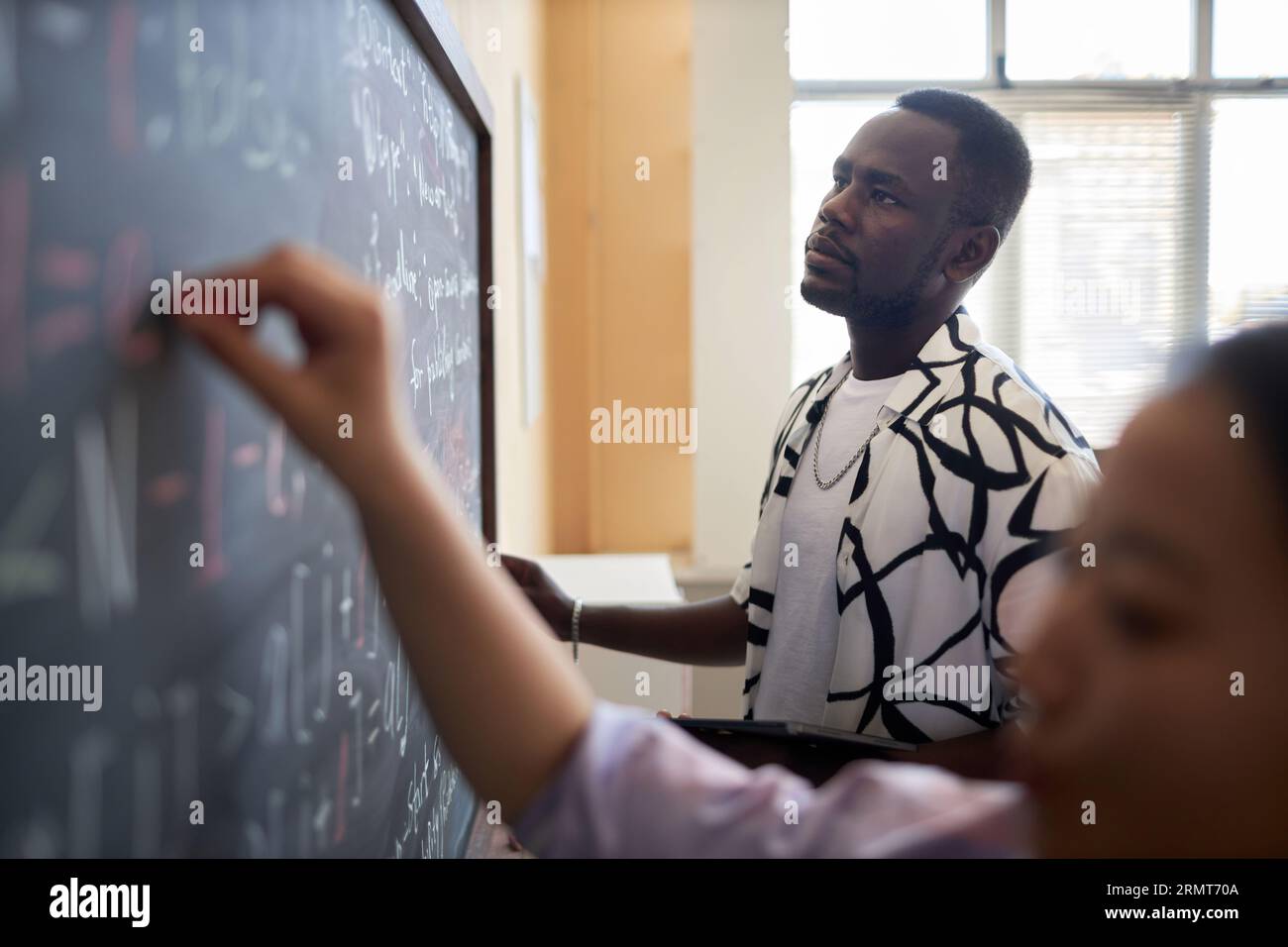 Young serious man in casualwear looking at equations on blackboard and ...