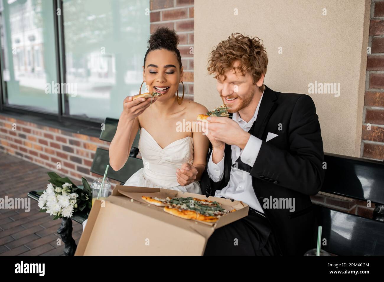 smiling african american bride and redhead groom eating pizza on bench ...