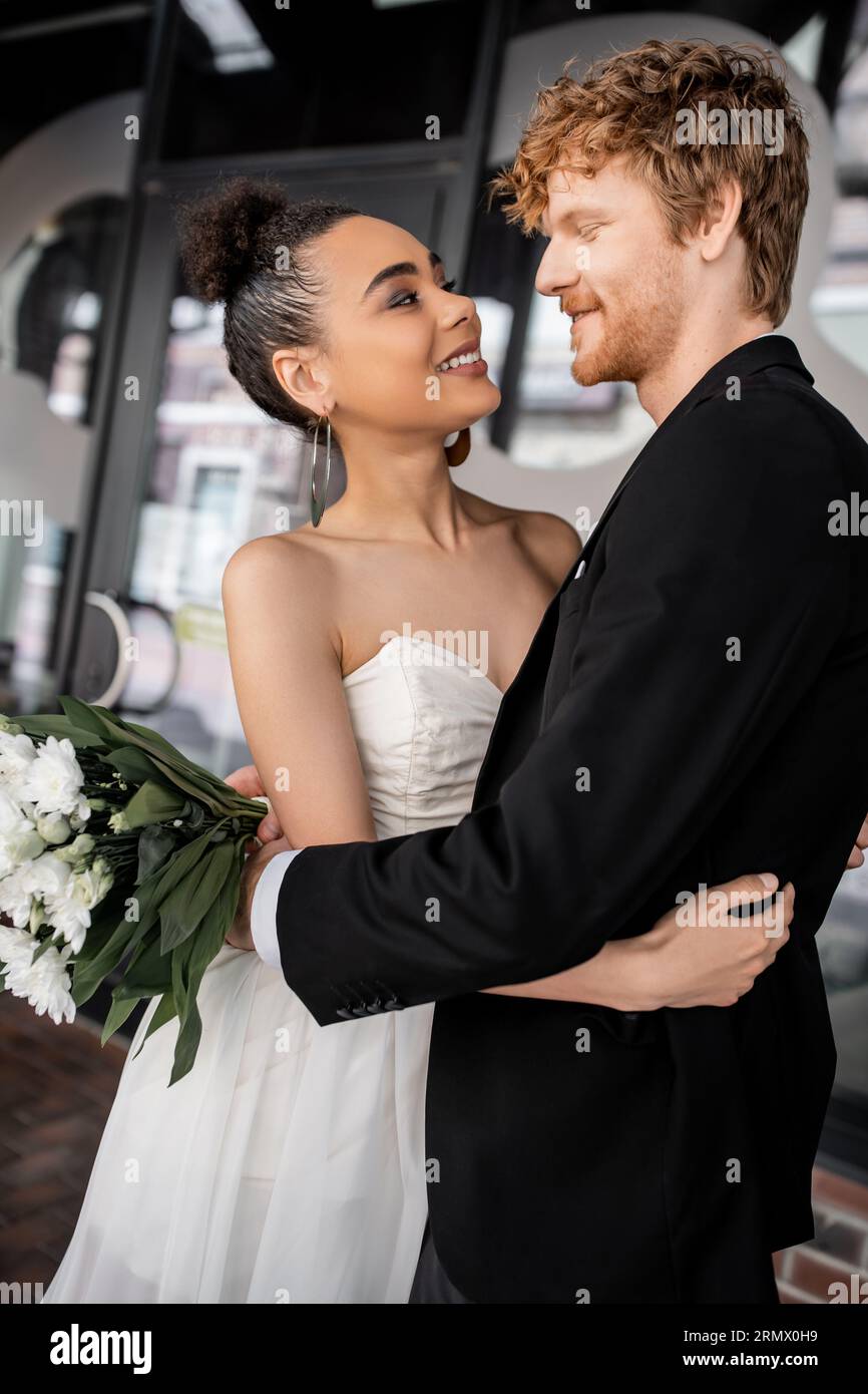 Overjoyed And Elegant Interracial Couple In Wedding Attire Embracing On