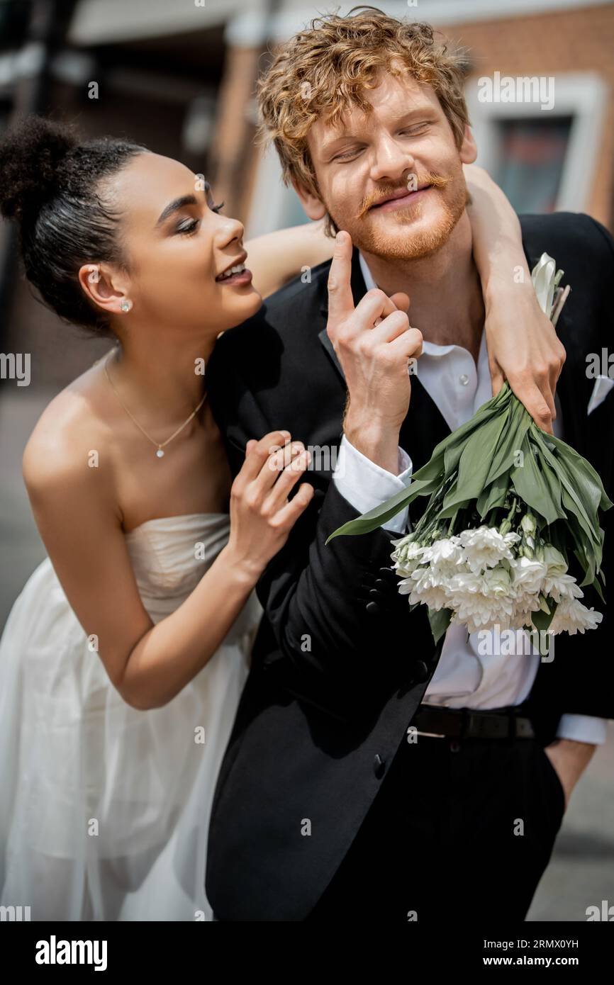 happy redhead man pointing at cheek near happy african american bride ...
