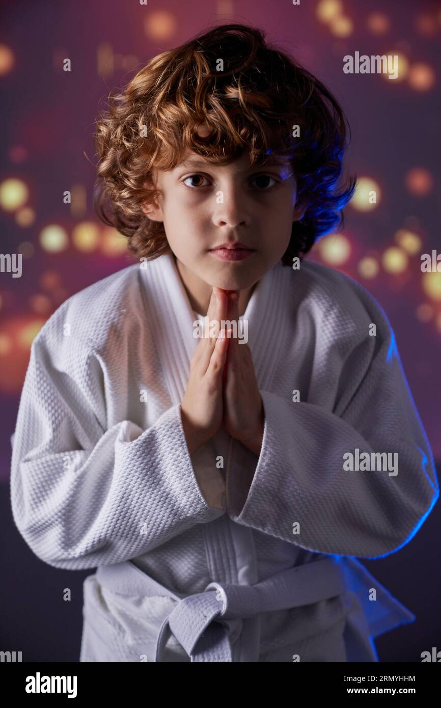 Cute kid in kimono looking at camera with folded hands while doing greeting gesture during judo ...