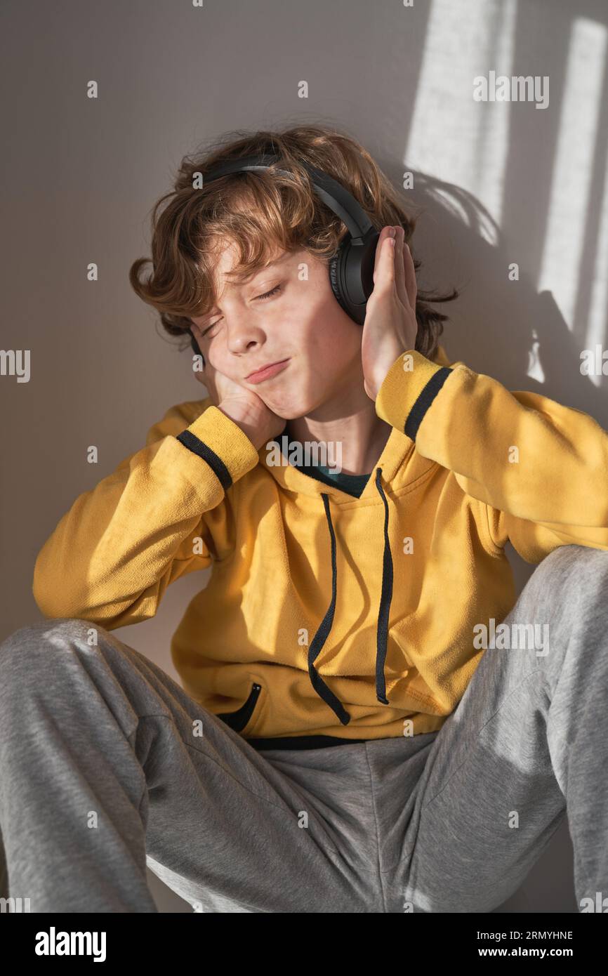 Pleased child in headphones and yellow sweater and gray pants sitting ...