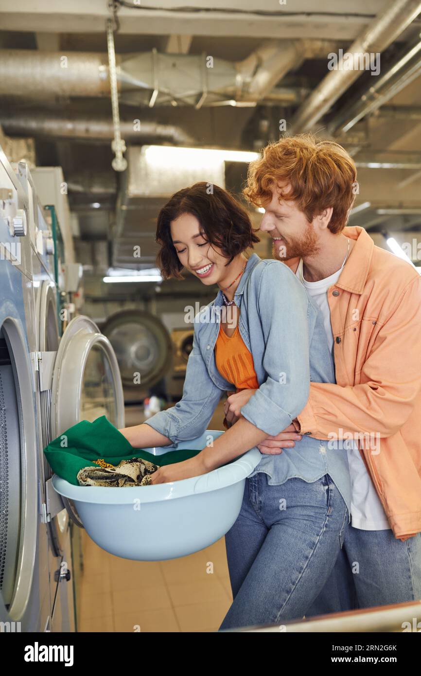 smiling young man hugging asian girlfriend with clothes near washing machine in public laundry ...