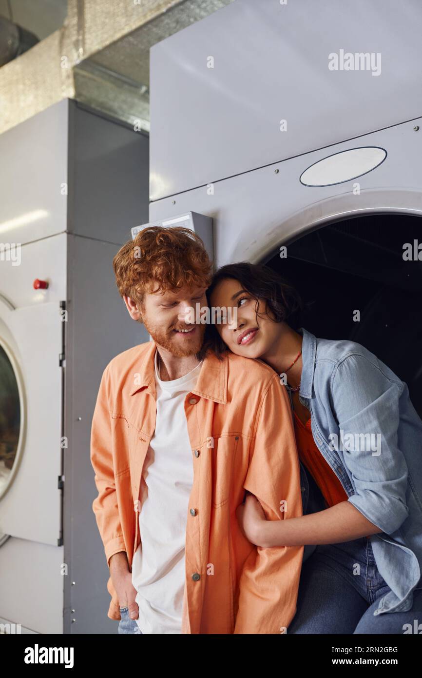 smiling young asian woman hugging and looking at boyfriend near washing machine in public ...