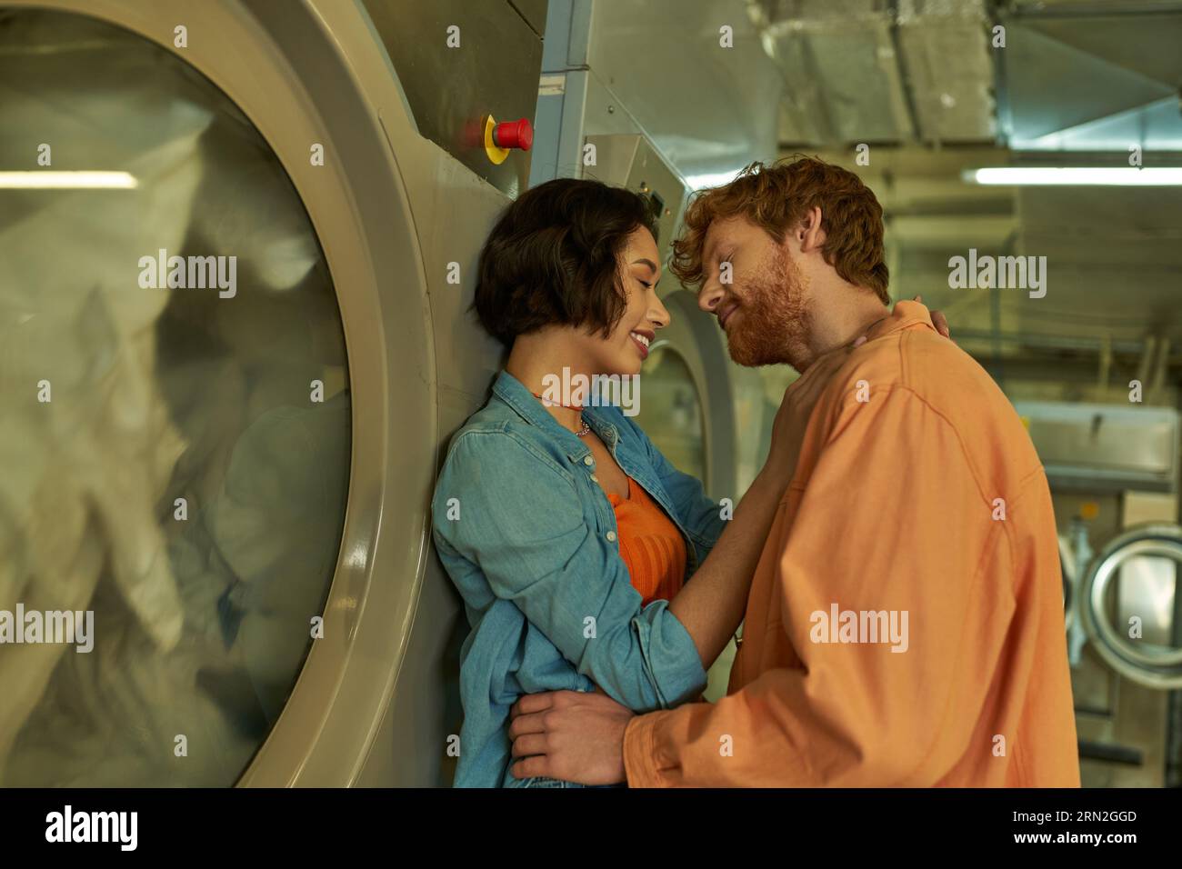 smiling multiethnic romantic couple kissing near washing machine in ...