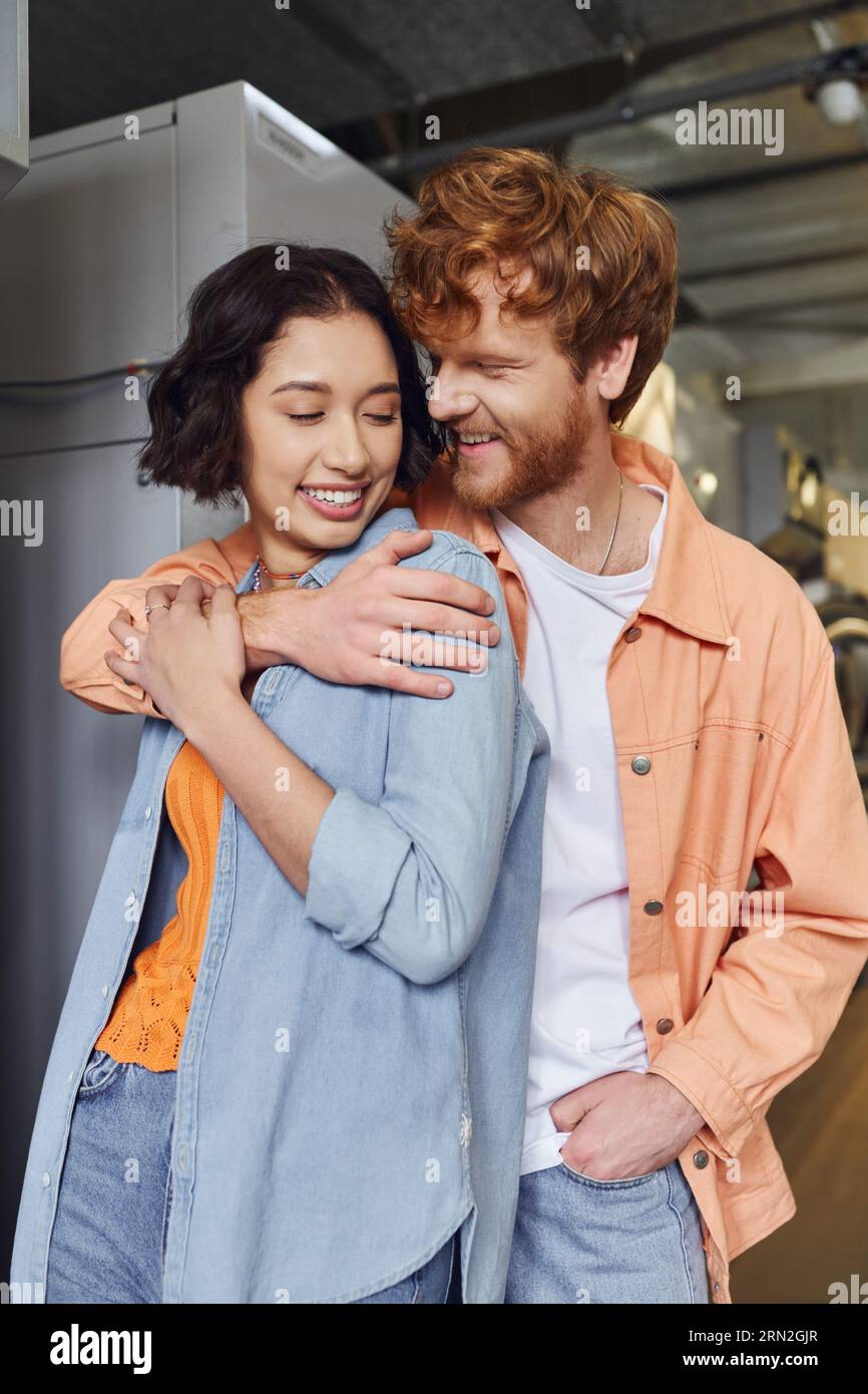 cheerful redhead man embracing brunette asian girlfriend while flirting ...