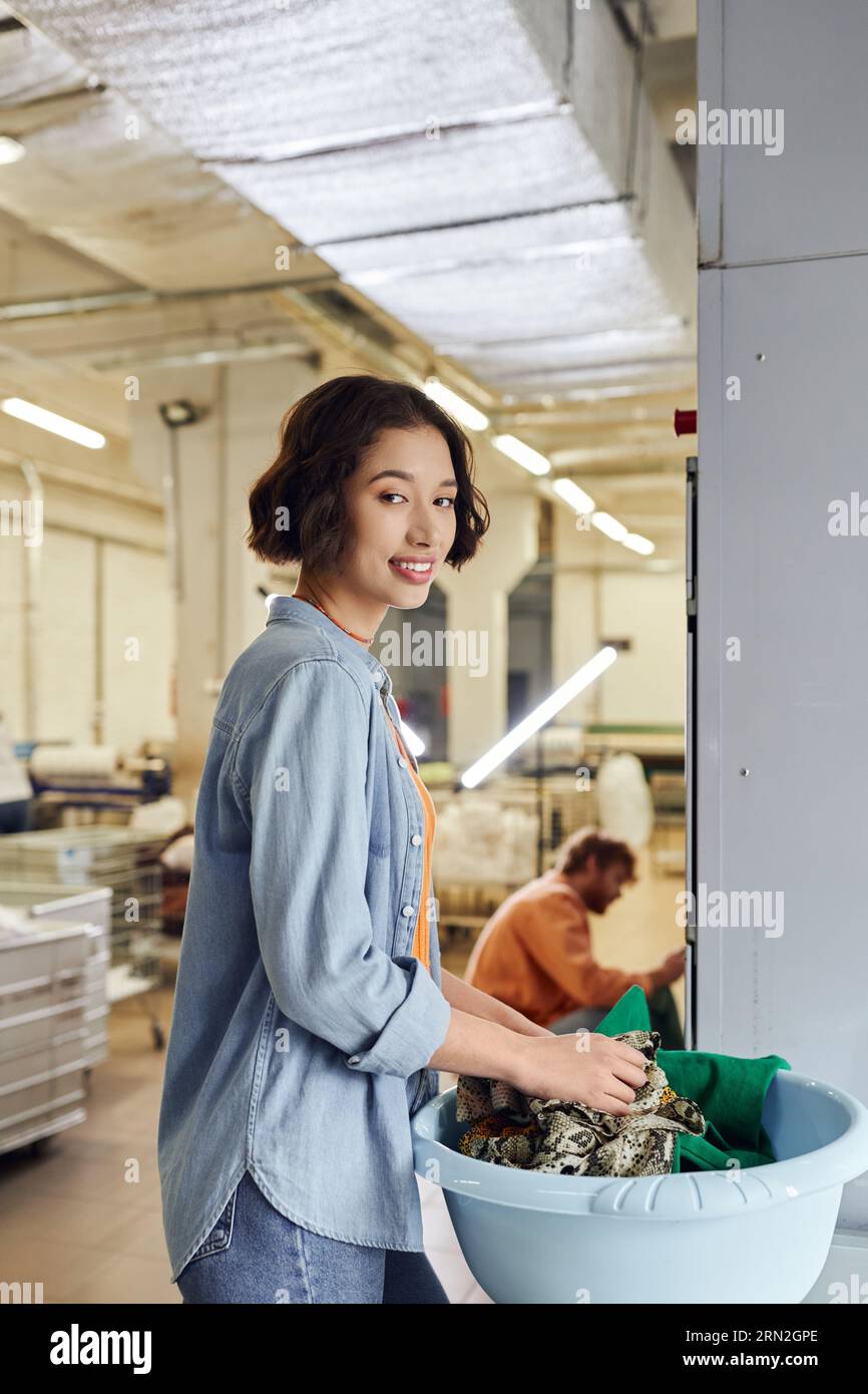 smiling young asian woman looking at camera near clothes in basket in blurred public laundry ...