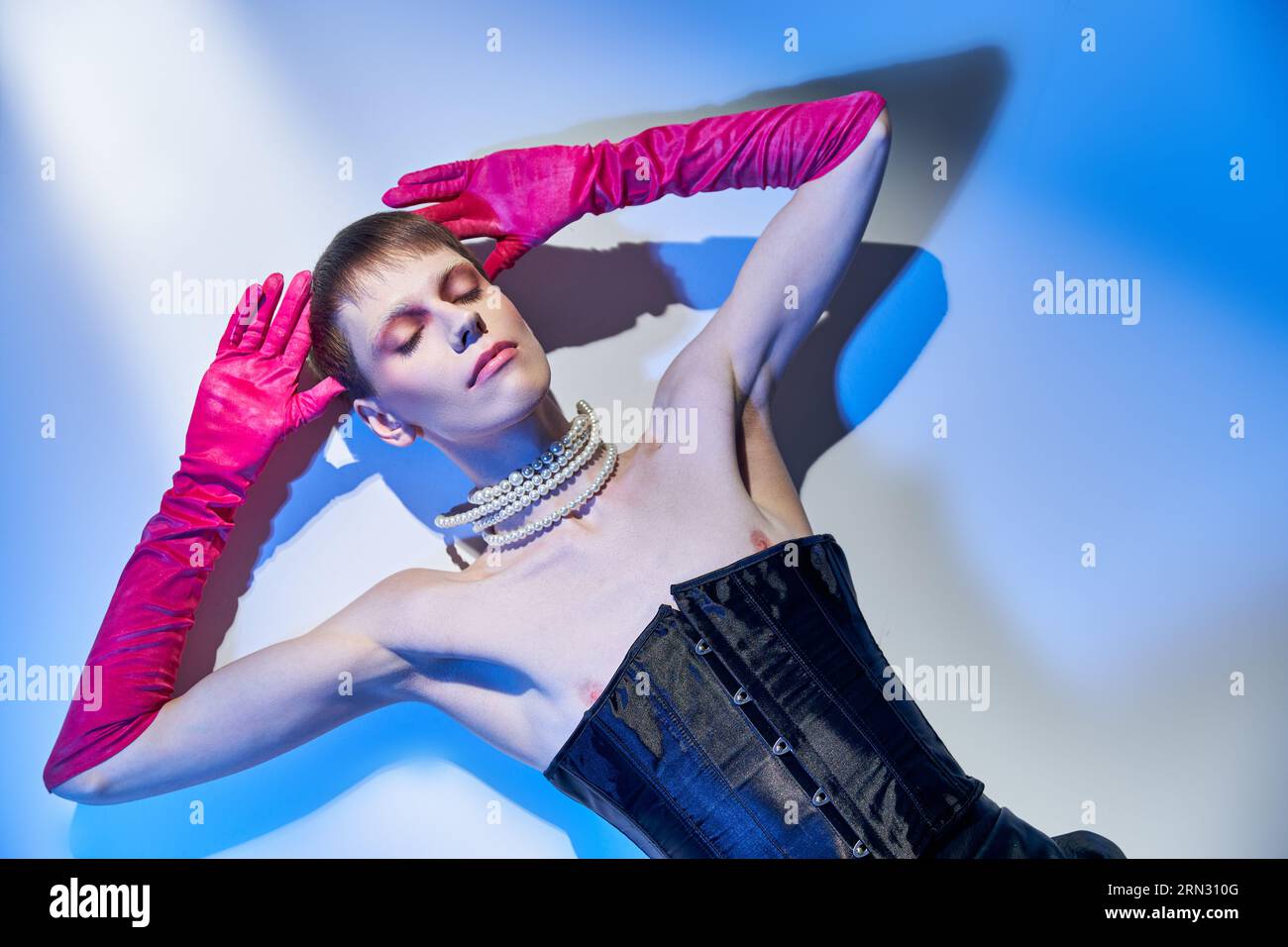 top view, queer model in corset and pink gloves lying on blue backdrop ...