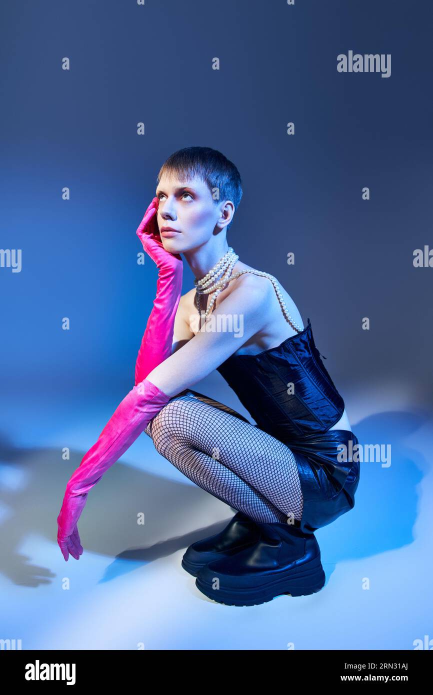 dreamy queer model in corset and shorts sitting on blue backdrop ...