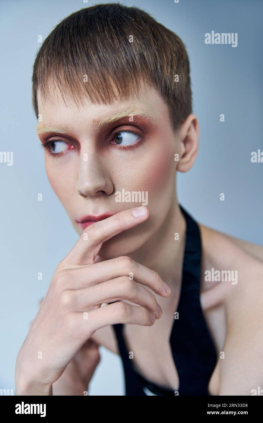 queer model with makeup looking away on grey backdrop, androgynous ...